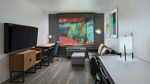 Quarto de hotel moderno com uma TV de tela plana grande, uma escrivaninha, um sofá confortável e uma janela ampla com uma pintura abstrata colorida.