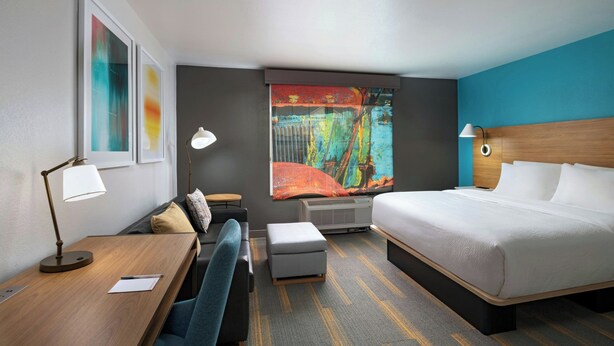 Quarto de hotel com cama, escrivaninha iluminada por abajur, poltrona, sofá e uma grande pintura abstrata na parede.