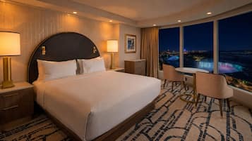 Quarto de hotel com uma cama grande, luminárias de cabeceira, uma escrivaninha, uma cadeira e vista para uma cascata à noite.