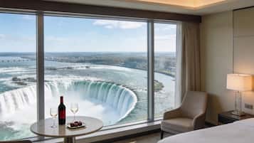 Quarto de hotel com uma grande janela com vista para uma cascata, uma cama, uma cadeira, uma mesa com uma garrafa e copos, e um abajur.