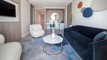 Uma sala de estar moderna com um sofá, uma poltrona branca, uma mesa de centro redonda e uma TV na parede.