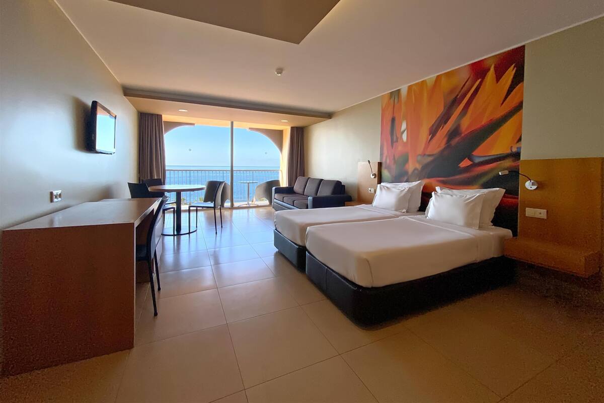 Quarto de hotel com varanda, escrivaninha, duas camas e um grande quadro abstrato.