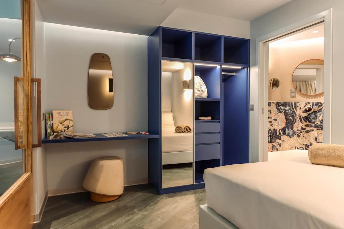 Quarto de hotel moderno com uma cama, uma escrivaninha com cadeira, um guarda-roupa e um espelho.