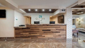 Uma área de recepção de hotel com piso de mármore, balcão de madeira e uma placa com a inscrição 'QUALITY INN'.