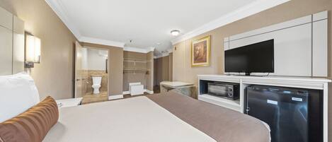 Quarto de hotel com uma cama grande, televisão, frigorífico e casa de banho visível através de uma porta aberta.