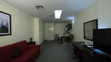 Quarto de hotel com sofá vermelho, escrivaninha com computador, televisão e cozinha compacta.