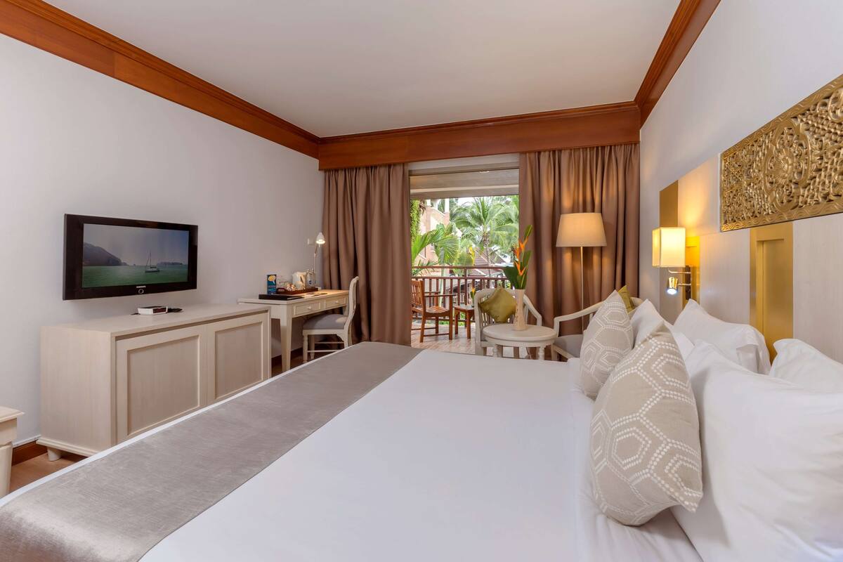 Quarto de hotel com uma cama grande, uma escrivaninha, uma televisão e uma varanda com cortinas.