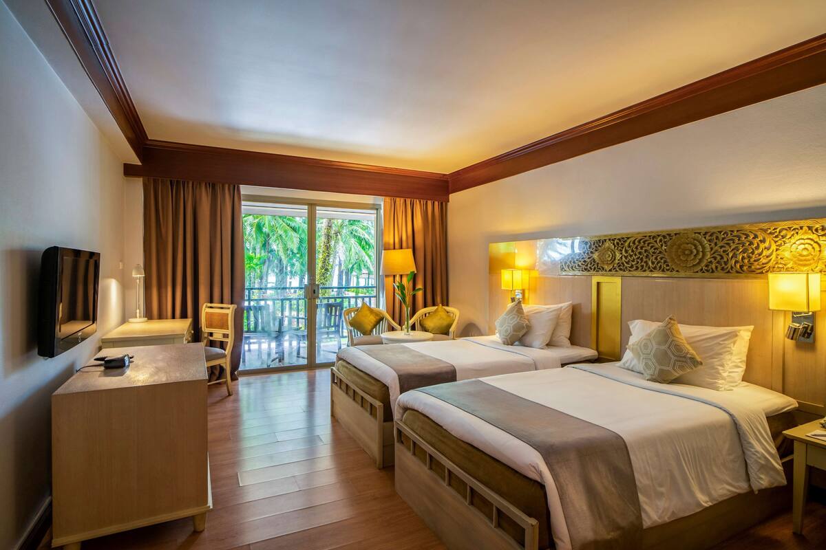 Quarto de hotel com duas camas, uma escrivaninha, televisão e varanda com vista para palmeiras.