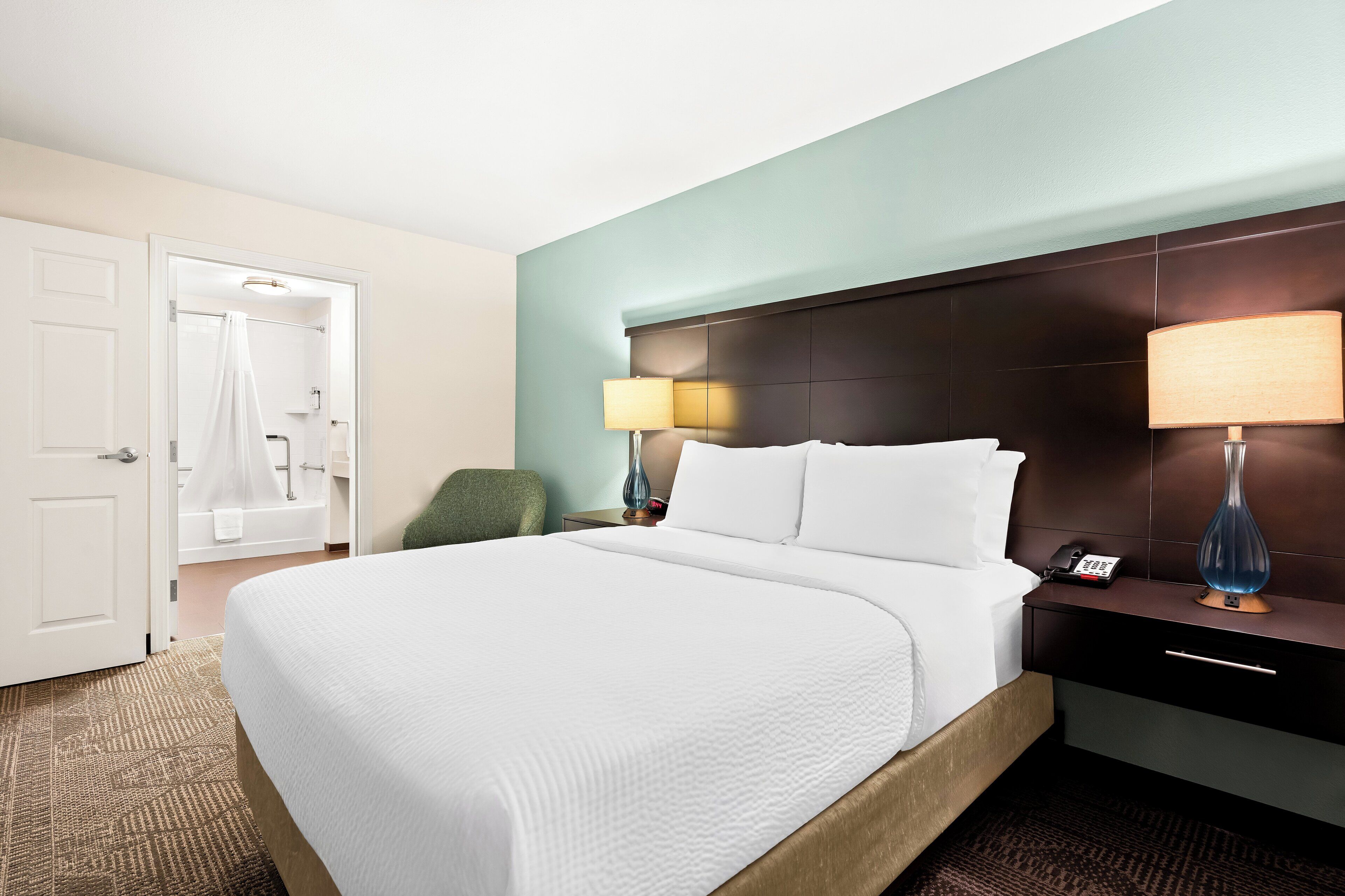 Quarto de hotel com cama grande, abajures, mesa de cabeceira e banheiro com chuveiro e vaso sanitário.