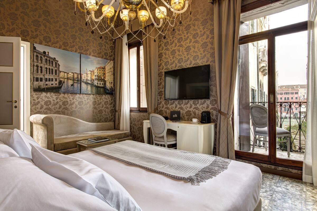 Quarto de hotel com cama, escrivaninha, cadeira, televisão e varanda com vista para edifícios.