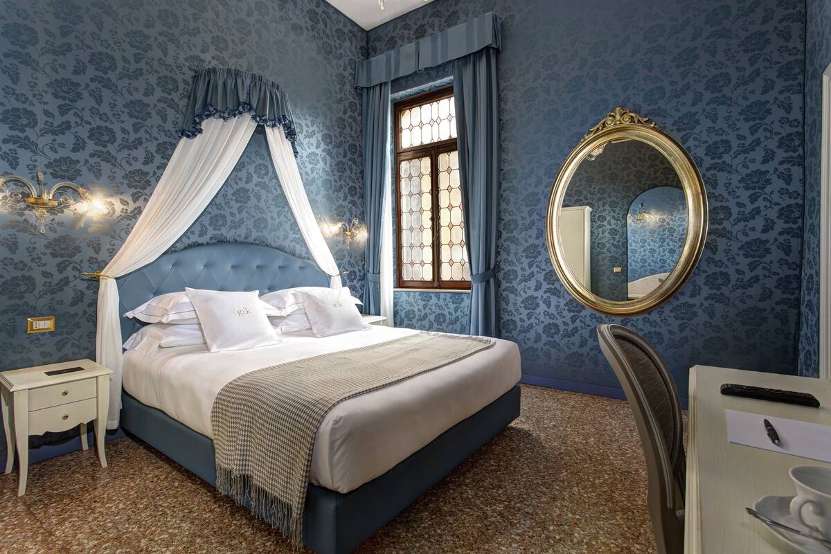 Quarto com cama, mesa de cabeceira, escrivaninha com cadeira, espelho e janela com cortinas.