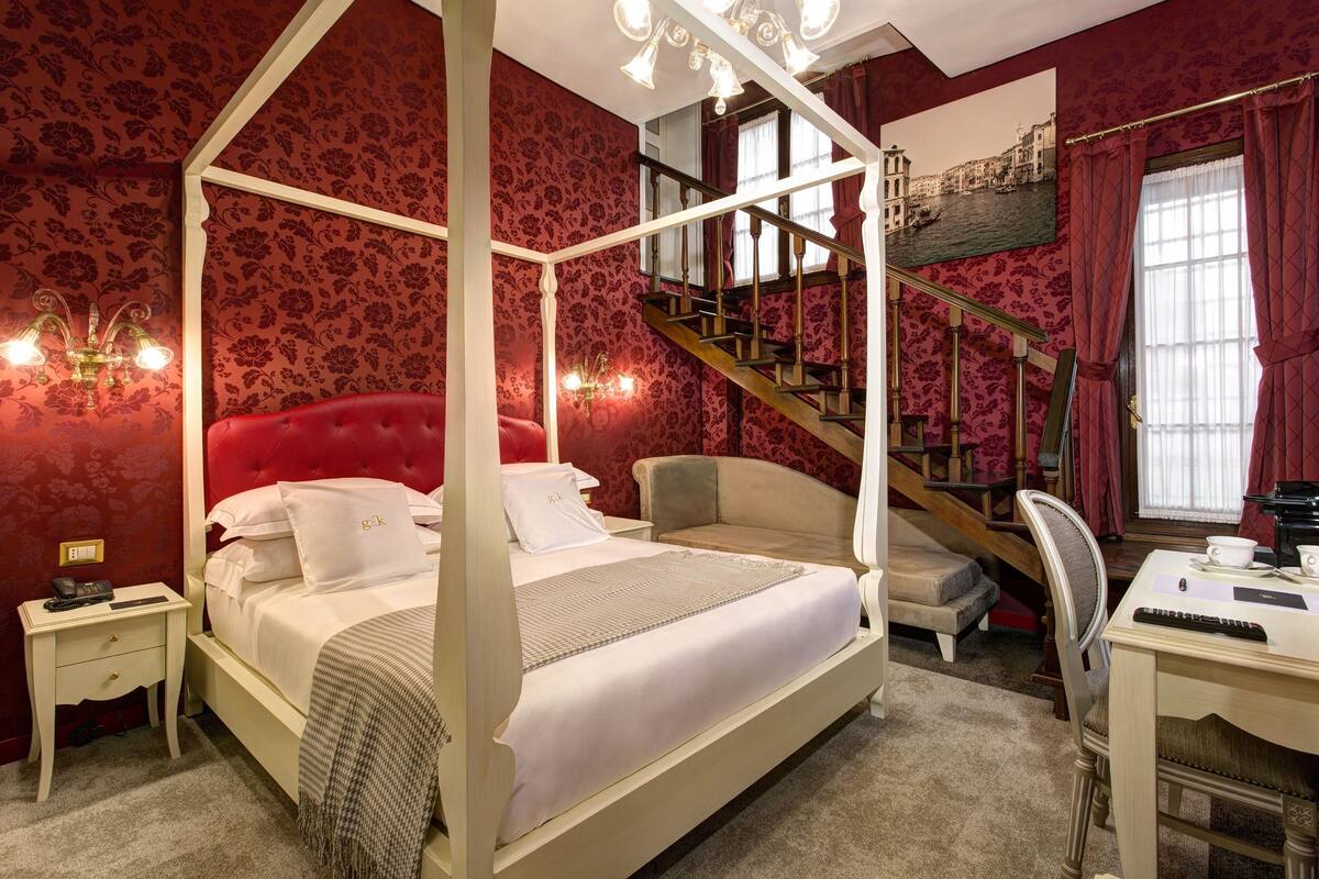 Uma cama com dossel e cabeceira vermelha, uma escrivaninha de madeira com cadeira, uma escada e uma parede com papel de parede floral.