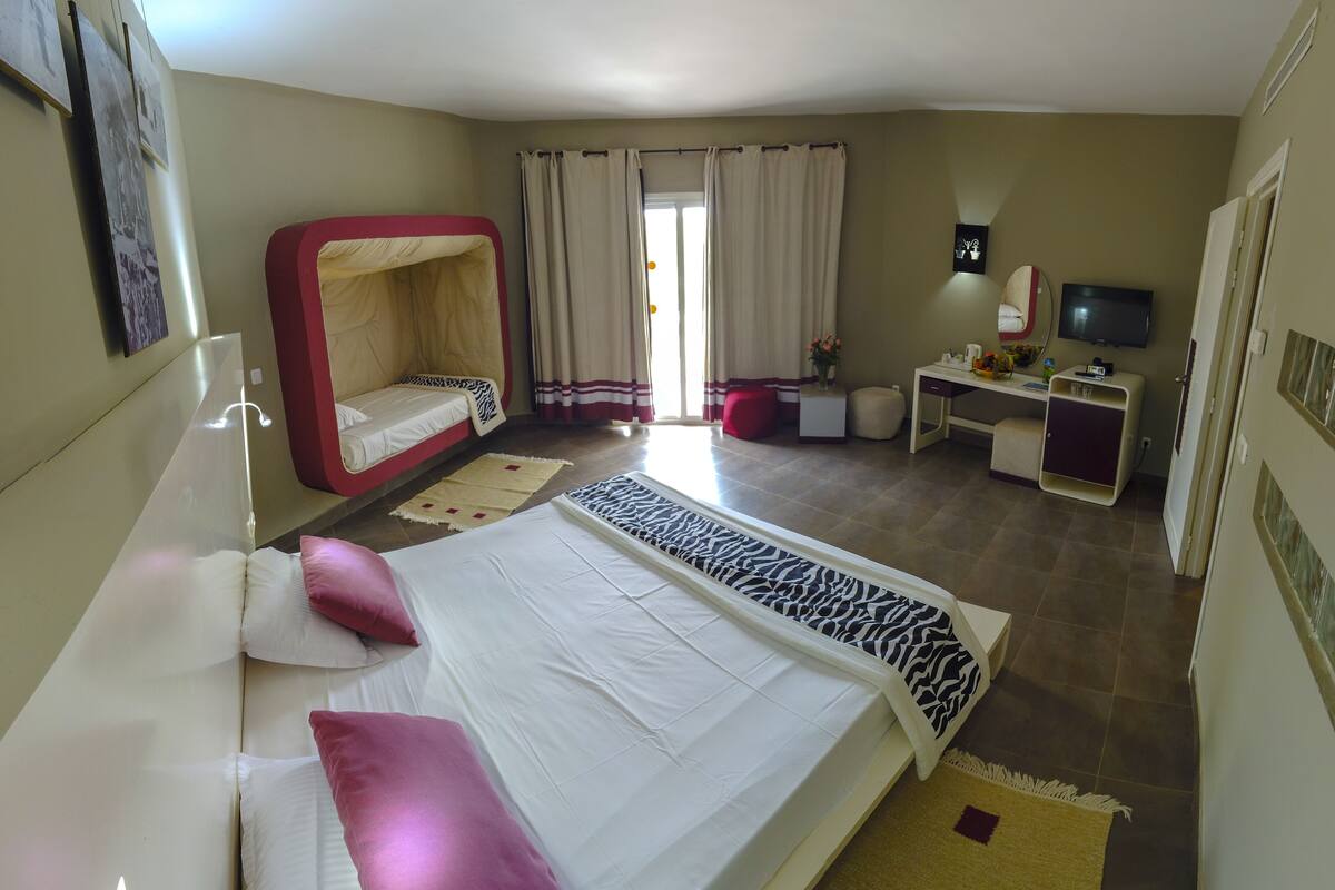 Quarto de hotel com uma cama grande, um sofá vermelho, uma escrivaninha com televisão e uma janela com cortinas.