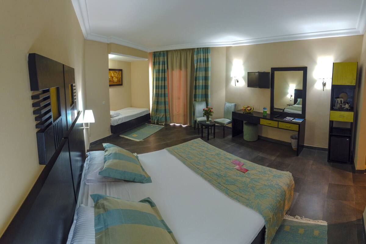 Quarto de hotel com uma cama grande, uma escrivaninha com cadeira, uma mesinha, um espelho e uma televisão.