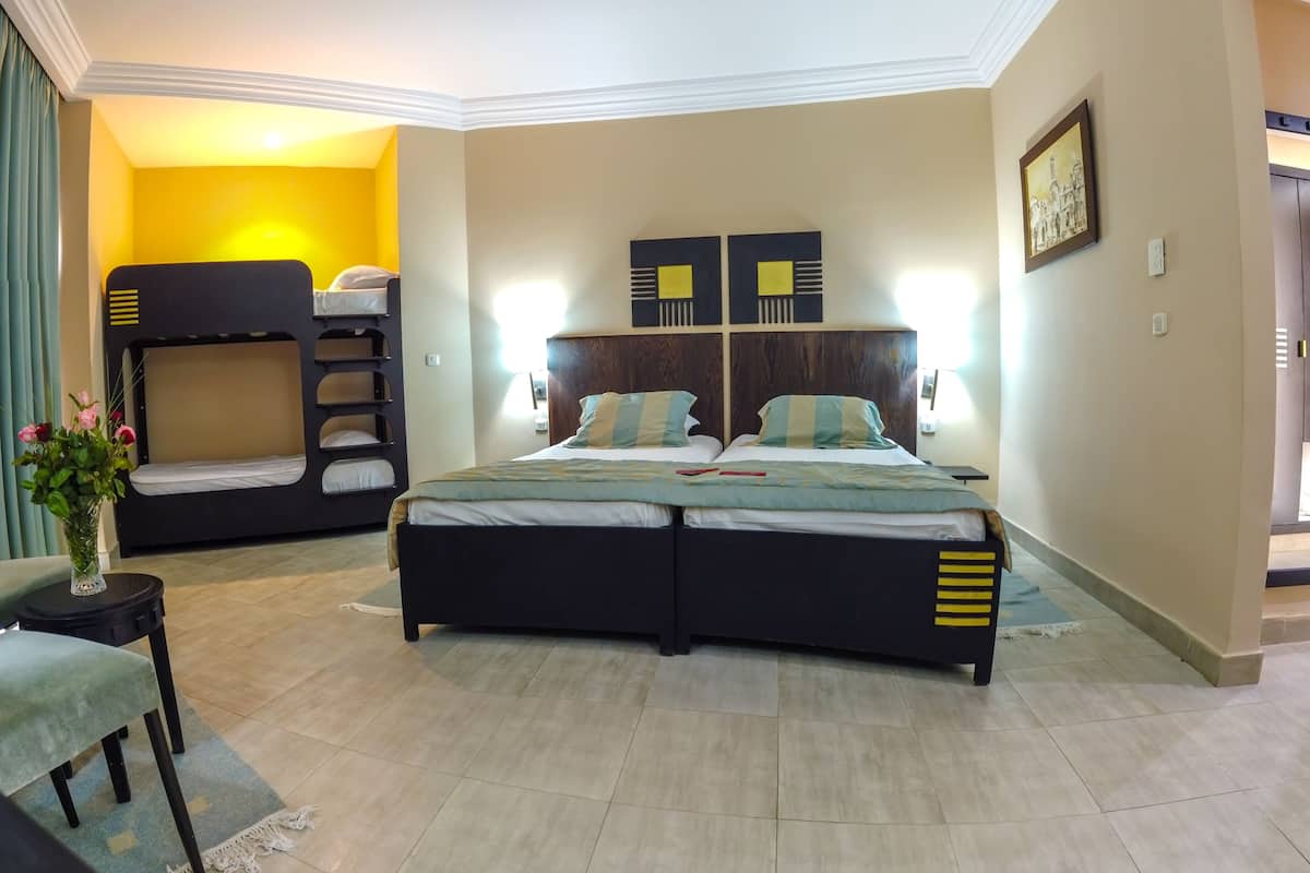 Quarto de hotel com beliche, uma cama de solteiro, uma cadeira, uma mesinha com um vaso de flores e um quadro na parede.