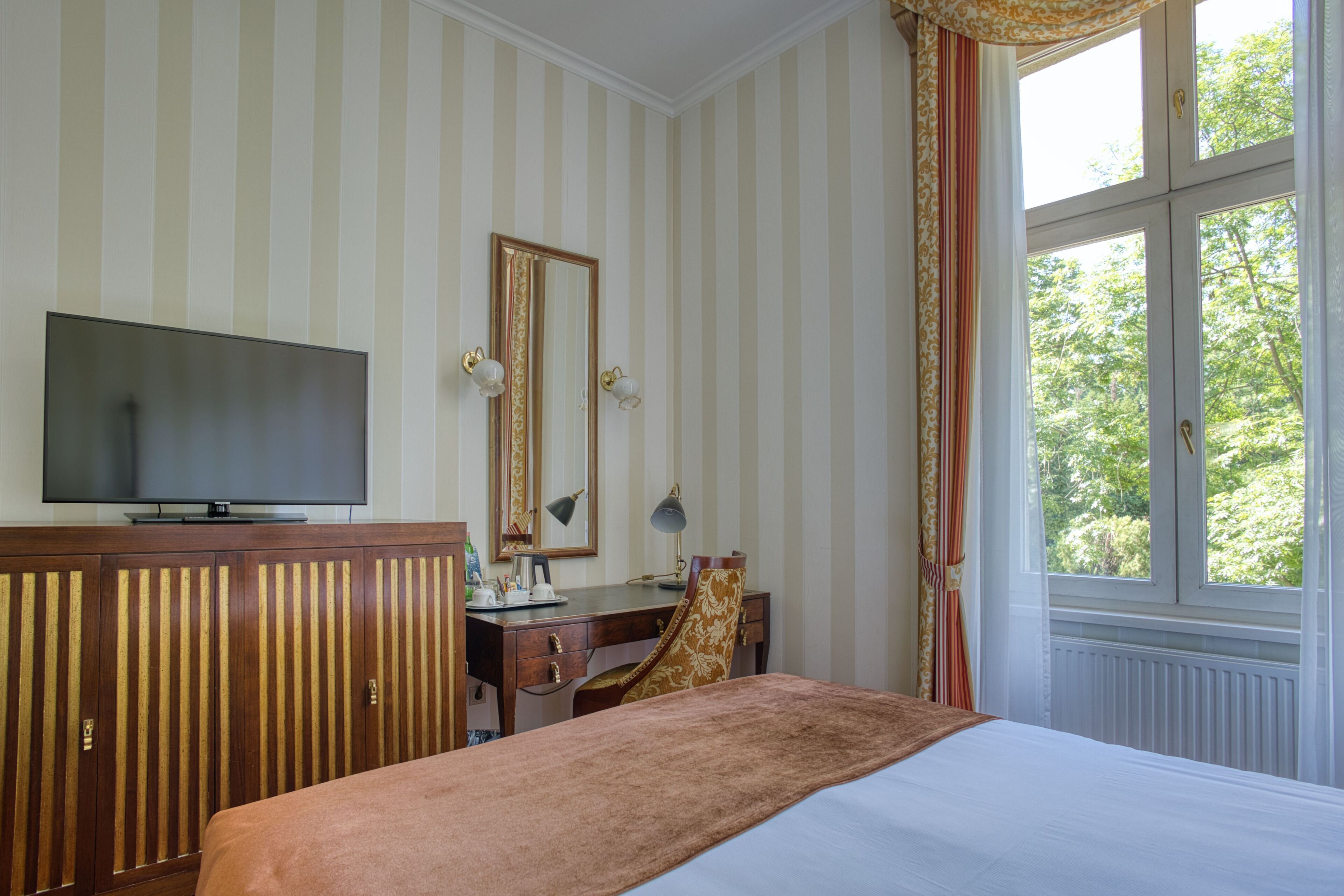 Quarto de hotel com cama, escrivaninha, televisão e janela com vista para árvores.