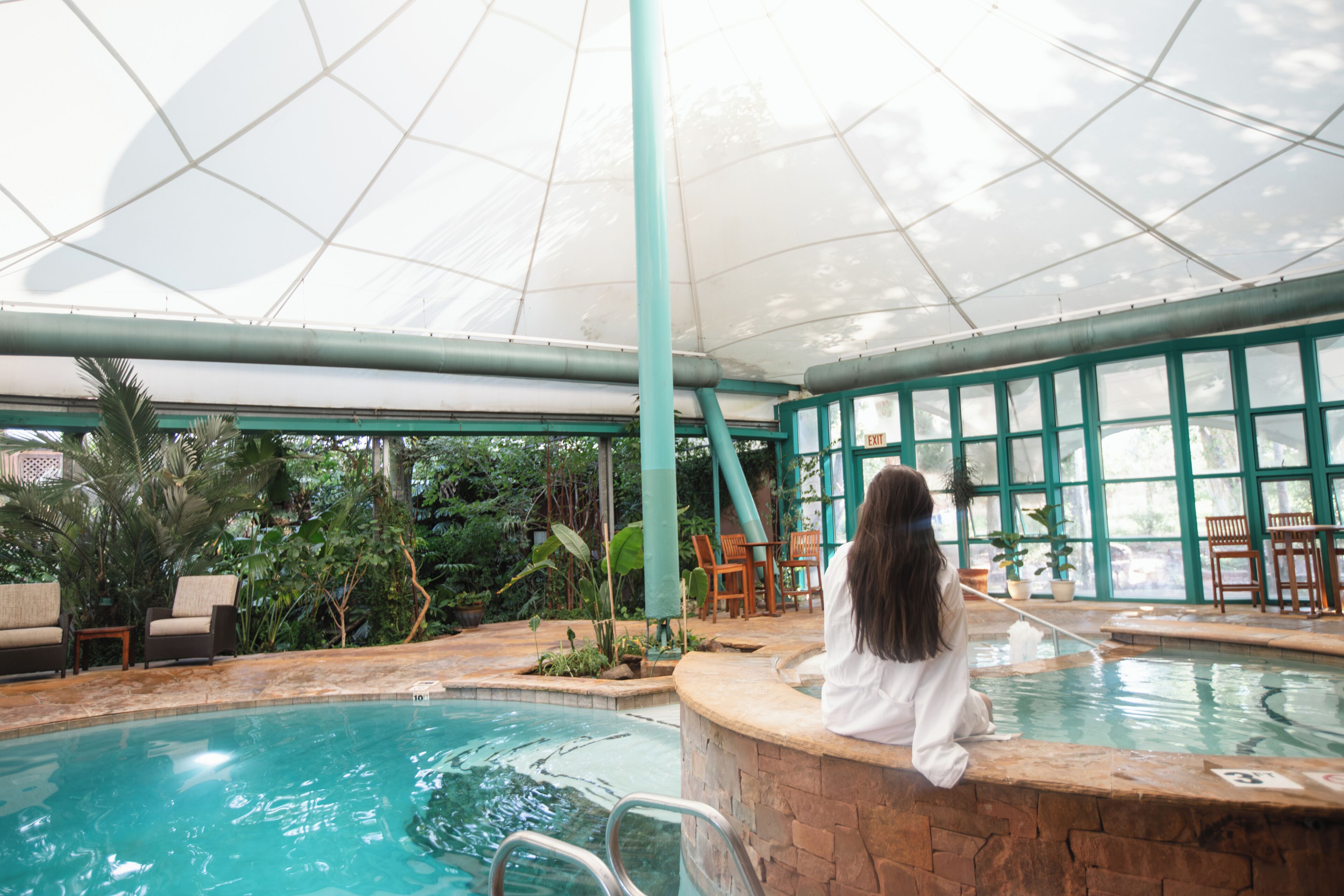 Piscina interna com teto de vidro, rodeada por vegetação e móveis de madeira.