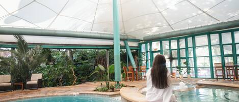 Piscina interna com teto de vidro, rodeada por vegetação e móveis de madeira.