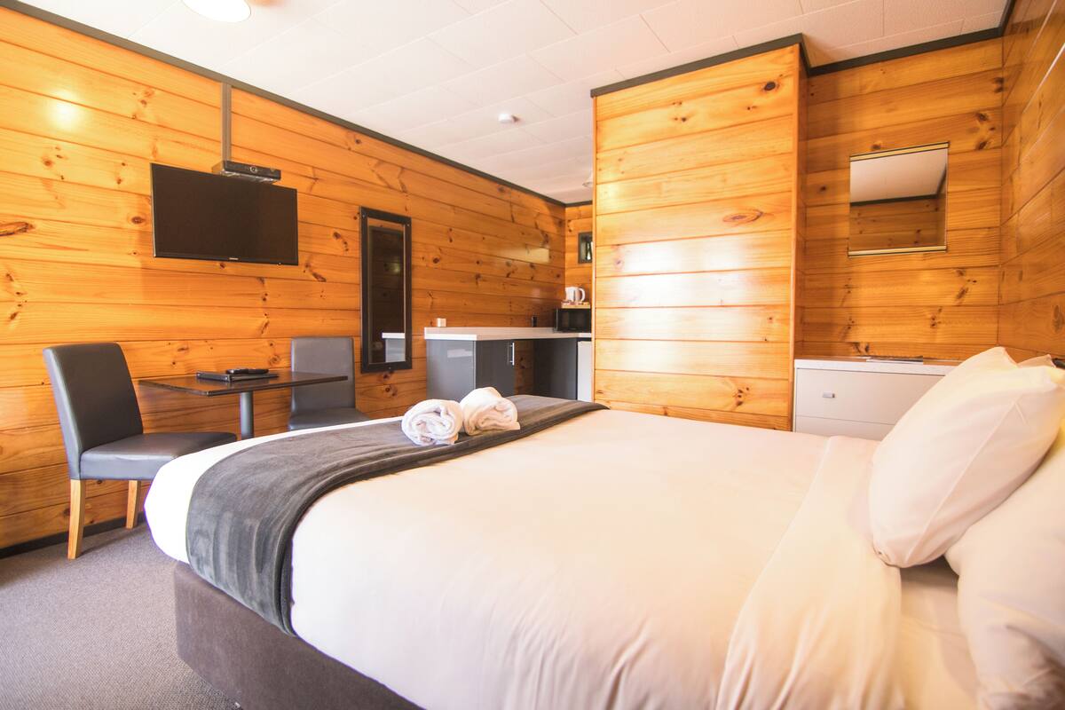 Quarto de hotel com uma cama, uma escrivaninha com cadeira, uma televisão fixada na parede e uma pequena área de kitchenette.