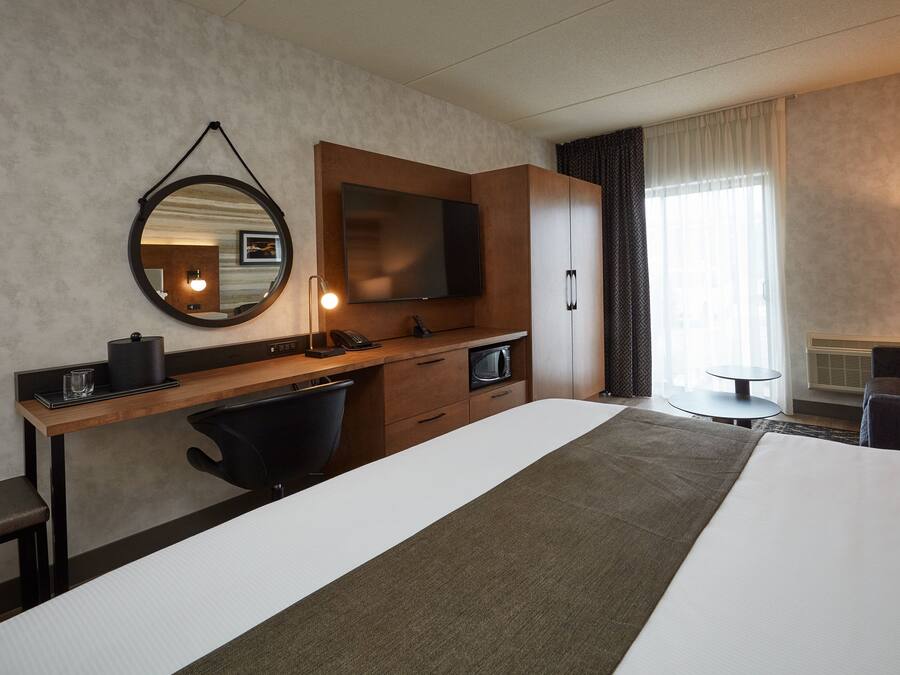 Quarto de hotel com cama, escrivaninha com computador, TV de tela plana, micro-ondas e pequena mesa com abajur.