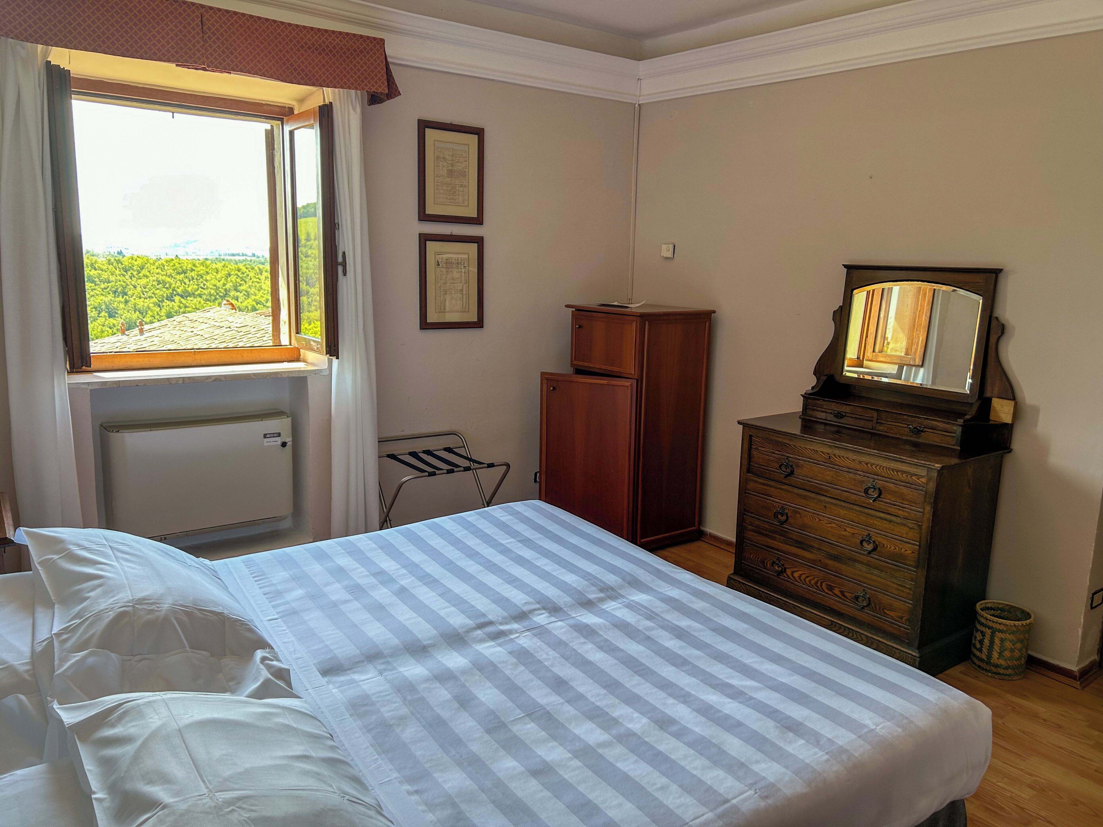 Quarto com cama, penteadeira, radiador, janela com cortinas e vista para o verde lá fora.