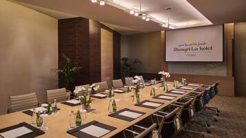 Uma sala de conferências no hotel Shangri-La em Dubai, com uma mesa longa, cadeiras e uma tela de projetor exibindo o nome do hotel.