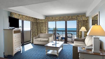 Quarto de hotel com sofá, poltrona, mesa de centro, TV e vista para o oceano.