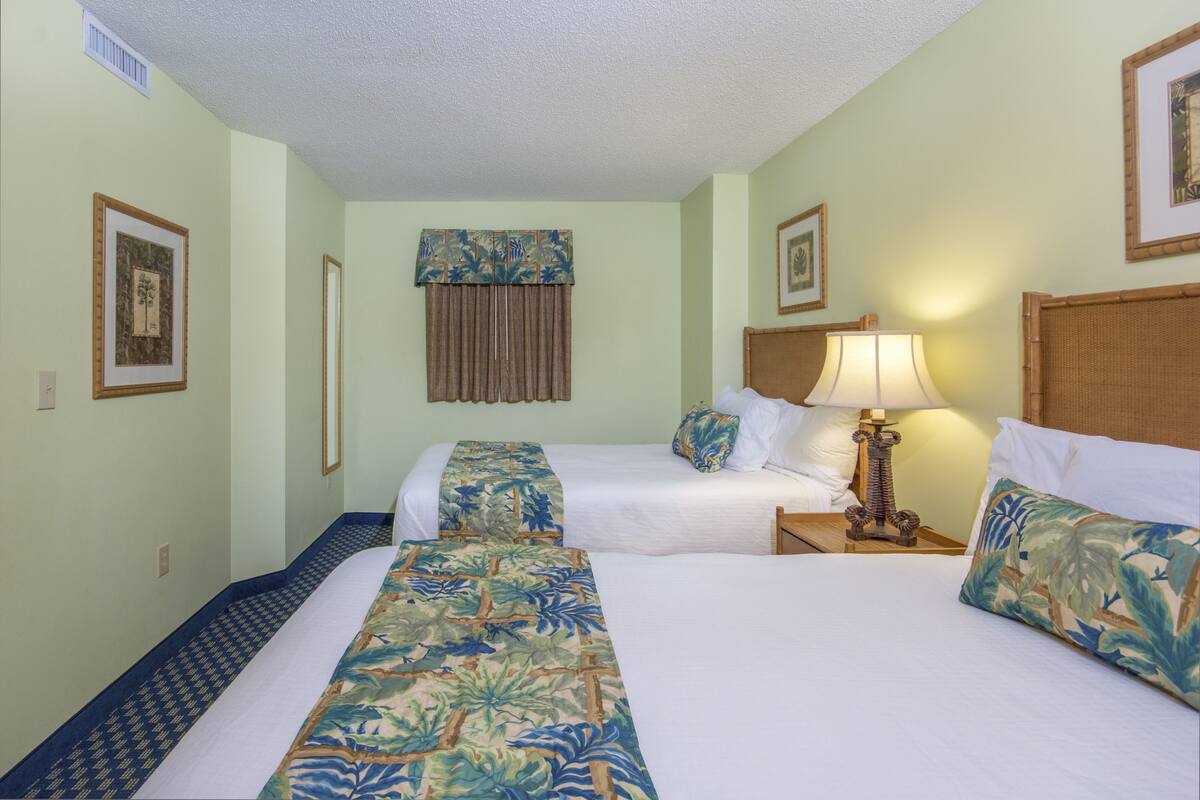 Quarto de hotel com duas camas, uma mesinha de cabeceira com abajur e uma janela com cortinas.