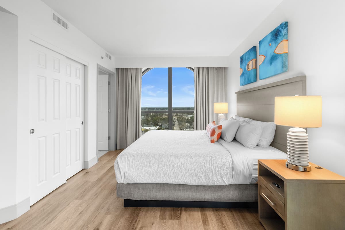 Quarto de hotel com uma cama grande, mesa de cabeceira com abajur e vista da cidade através de uma janela ampla.