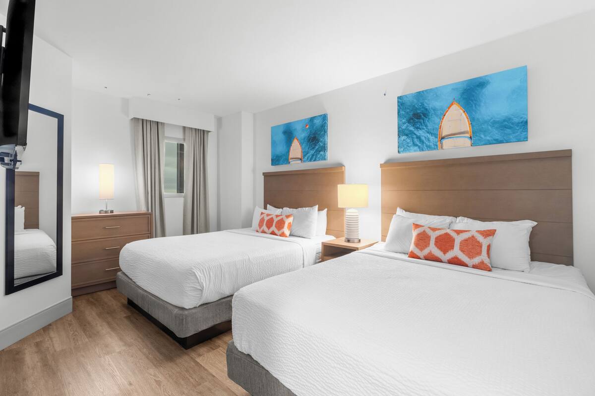 Quarto de hotel com duas camas, cabeceira de madeira, mesa de cabeceira com abajur, espelho e janela com cortinas.