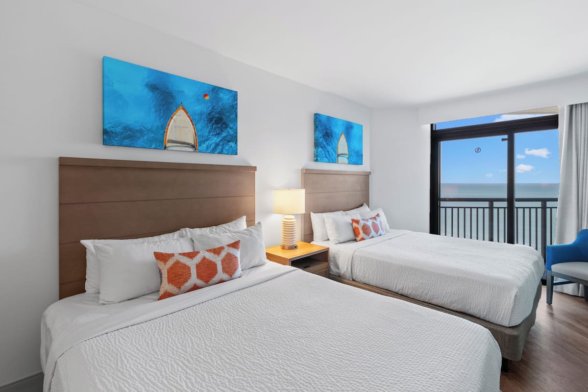 Quarto de hotel com duas camas, uma mesa de cabeceira com abajur e obras de arte nas paredes.