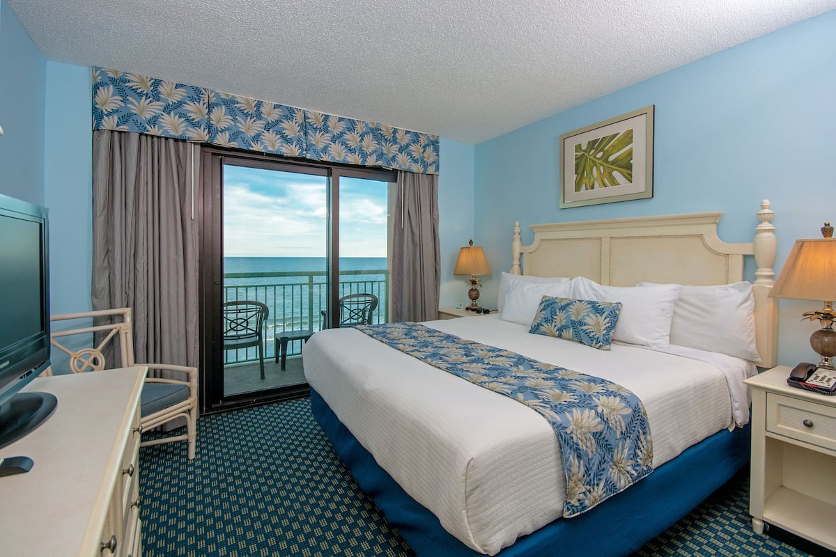 Quarto de hotel com uma cama grande, uma escrivaninha, uma cadeira, mesa de cabeceira com abajur e vista para o oceano através de uma porta deslizante.