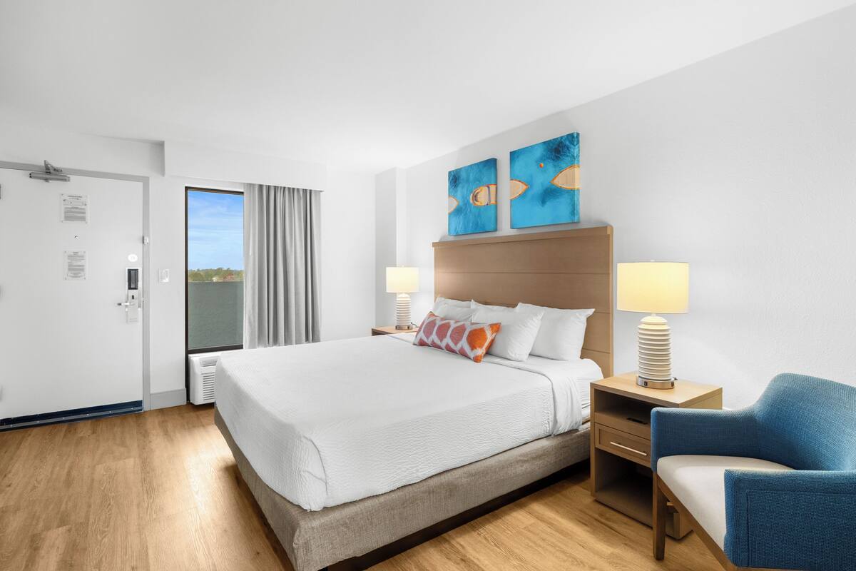 Quarto de hotel com uma cama grande, duas mesas de cabeceira com luminárias, uma cadeira e duas obras de arte abstratas nas paredes.