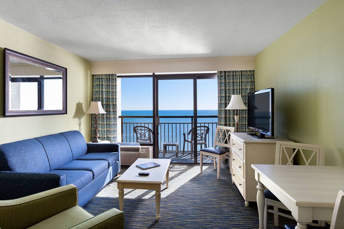 Quarto de hotel com varanda, sofá, escrivaninha, TV e vista para o oceano.