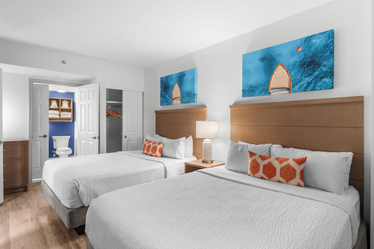 Quarto de hotel com duas camas, cabeceira de madeira, mesa de cabeceira com abajur e obras de arte nas paredes.
