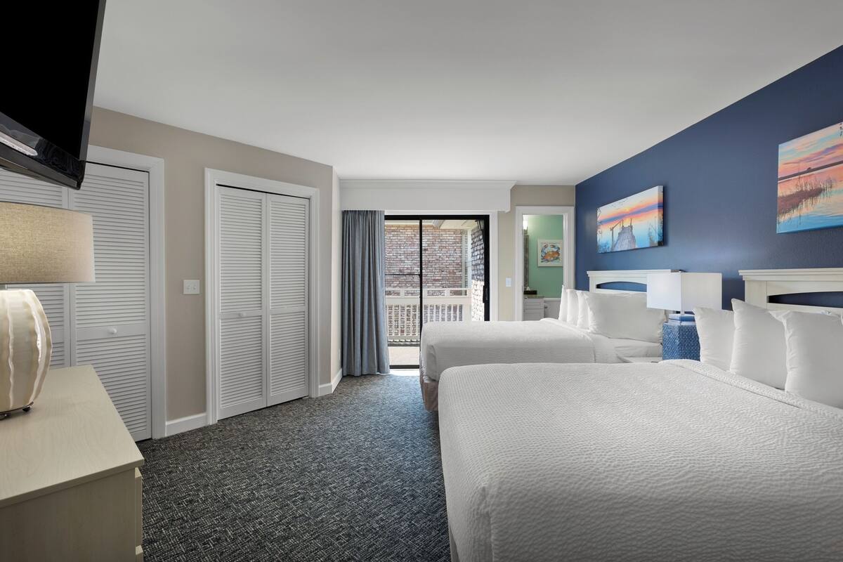 Quarto de hotel com duas camas, televisão, escrivaninha, janela com vista para uma varanda e obras de arte na parede.