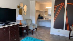 Quarto de hotel com televisão de tela plana, abajur, cadeira, escrivaninha e tapete estampado.