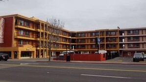 Um motel de vários andares com fachada em vermelho e amarelo, várias varandas e um estacionamento em frente.