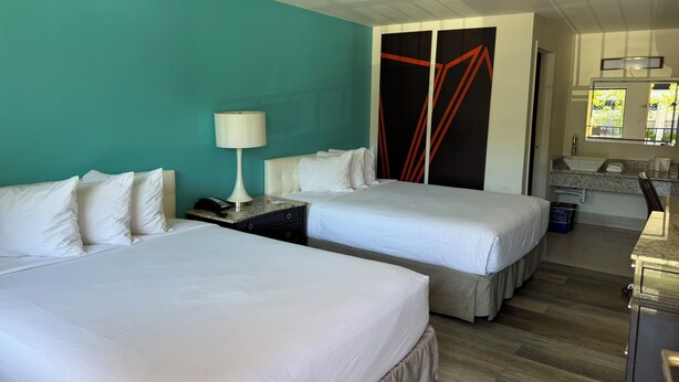 Quarto de hotel com duas camas, uma escrivaninha com luminária, banheiro com pia e espelho, e parede verde-azulado.