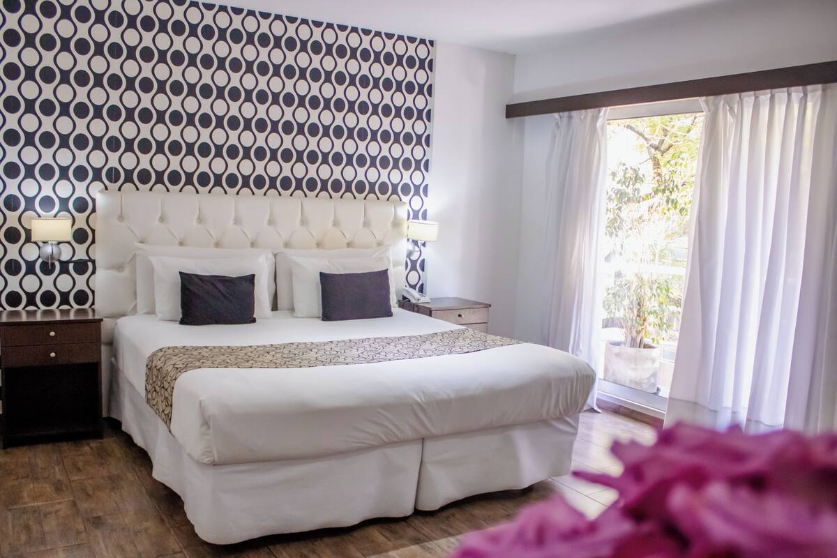 Quarto de hotel com uma cama grande, parede estampada, mesa de cabeceira e vista para o exterior.