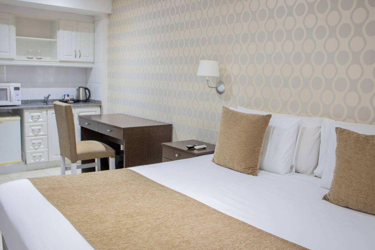 Quarto de hotel com cama, escrivaninha, cadeira, mesa de cabeceira e micro-ondas na área da cozinha.