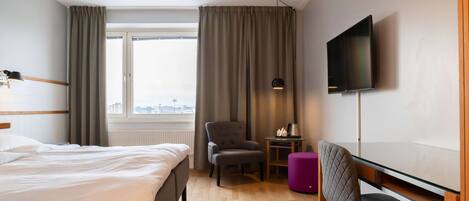 Quarto de hotel com cama, poltrona, mesinha de cabeceira com abajur, televisão e janela com cortinas.