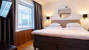 Quarto de hotel com uma cama grande, uma TV fixada na parede, uma janela com cortinas e duas luminárias.