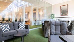 Um espaço interior moderno com parede de vidro, sofá com almofadas estampadas, cadeiras de vime e uma pequena mesa.