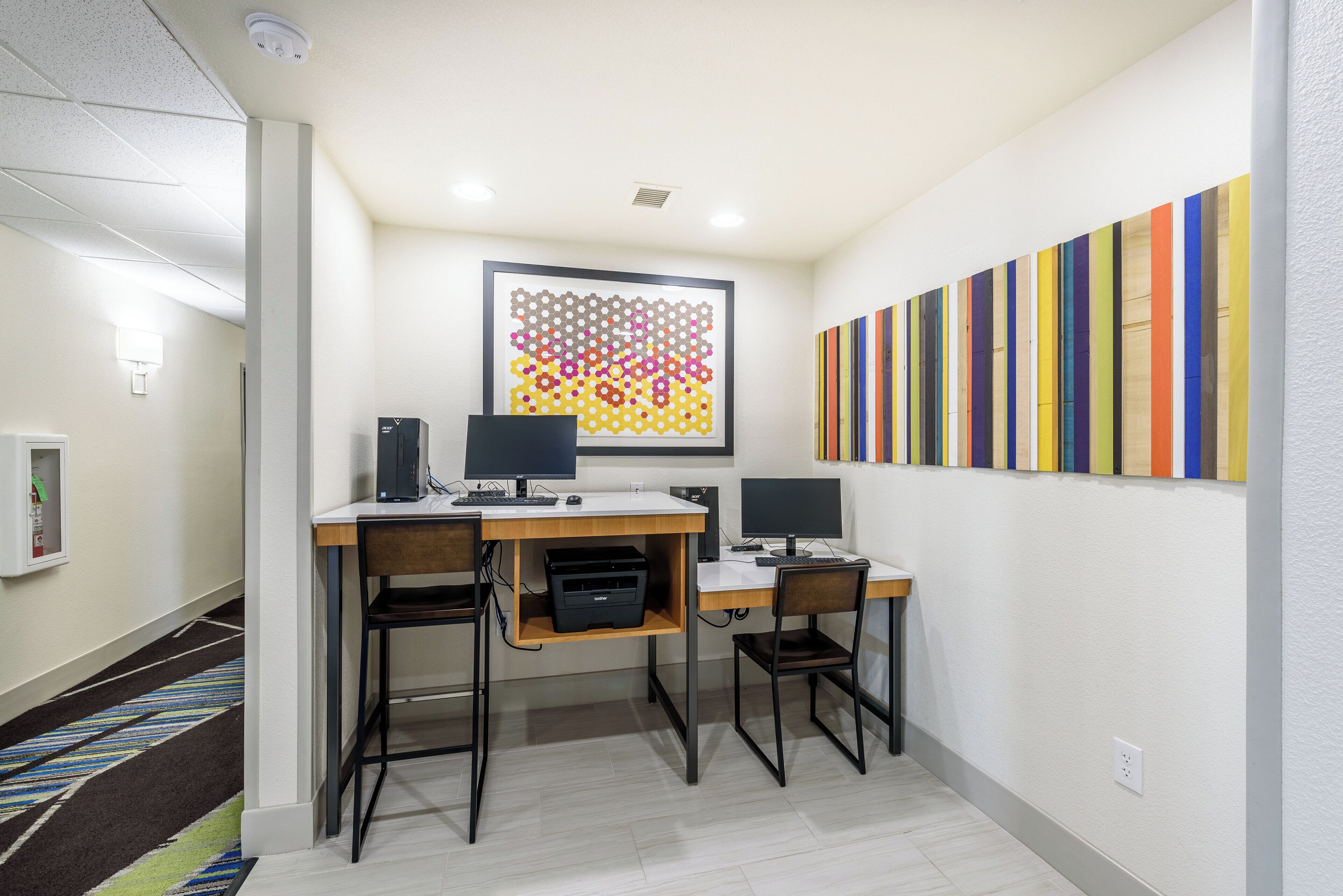 Espaço de escritório moderno com duas mesas, cada uma com um computador e uma impressora. Há uma obra de arte abstrata emoldurada na parede acima das mesas.
