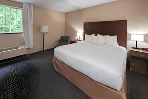 Quarto de hotel com uma cama grande, uma cadeira, uma escrivaninha e uma janela com cortinas.