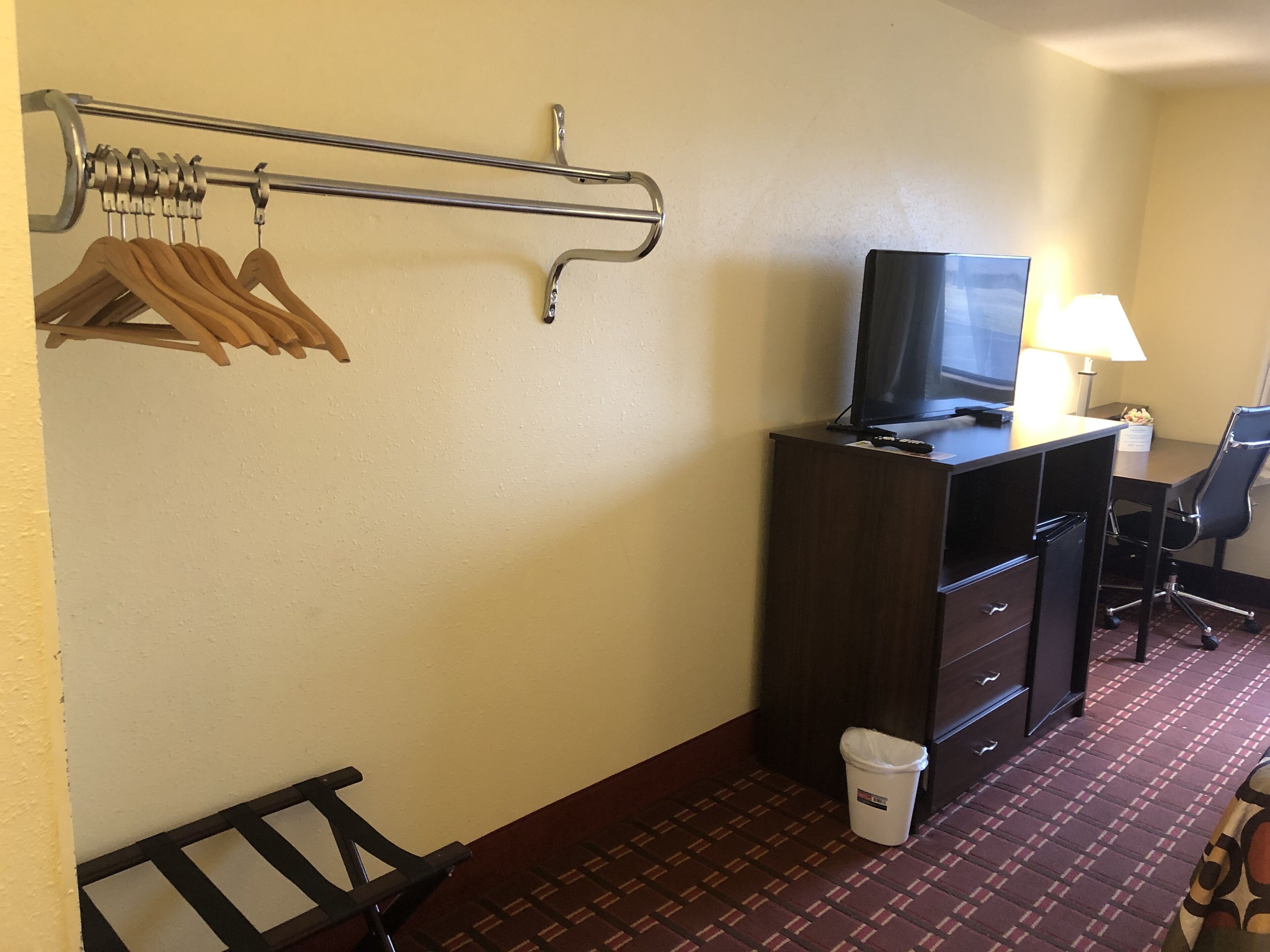 Quarto de hotel com um cabideiro de madeira, uma televisão sobre uma escrivaninha, uma luminária de mesa, uma cadeira de escritório preta, uma lixeira e um tapete estampado.