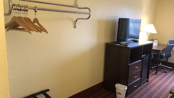 Quarto de hotel com um cabideiro de madeira, uma televisão sobre uma escrivaninha, uma luminária de mesa, uma cadeira de escritório preta, uma lixeira e um tapete estampado.