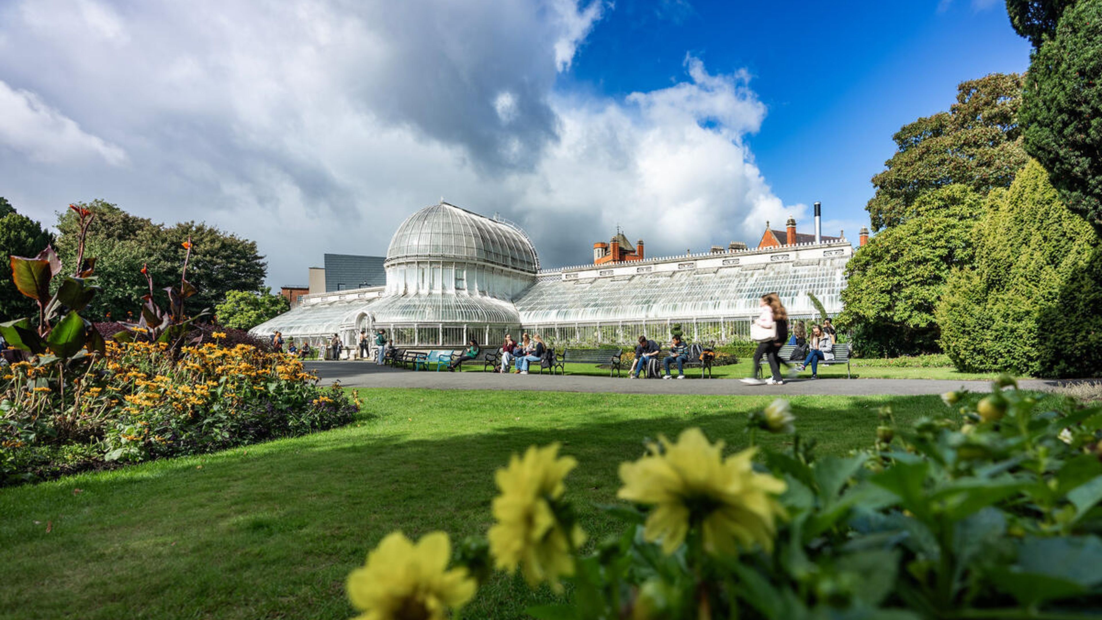 Uma grande estufa de vidro com estrutura em cúpula, rodeada por um jardim bem cuidado com diversas plantas e flores.