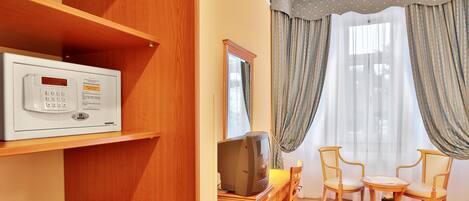 Quarto de hotel com um armário de madeira, uma televisão, uma mesa pequena com duas cadeiras e uma janela com cortinas.